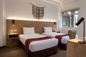 Quarto de hotel com duas camas, uma escrivaninha, uma cadeira, uma luminária na parede e uma janela.