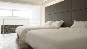 Quarto de hotel moderno com duas camas, uma janela grande e uma cabeceira escura.