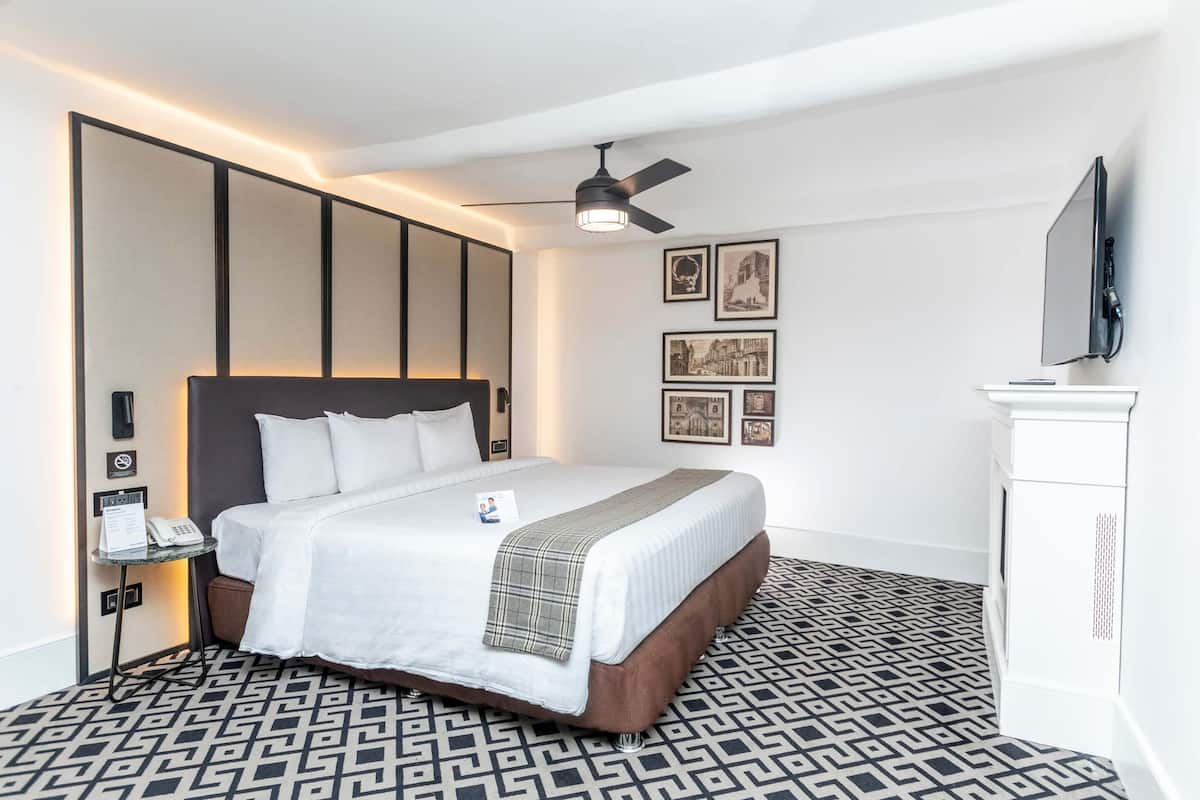 Quarto de hotel com uma cama grande, um ventilador de teto, uma mesa de cabeceira com telefone e quadros nas paredes.