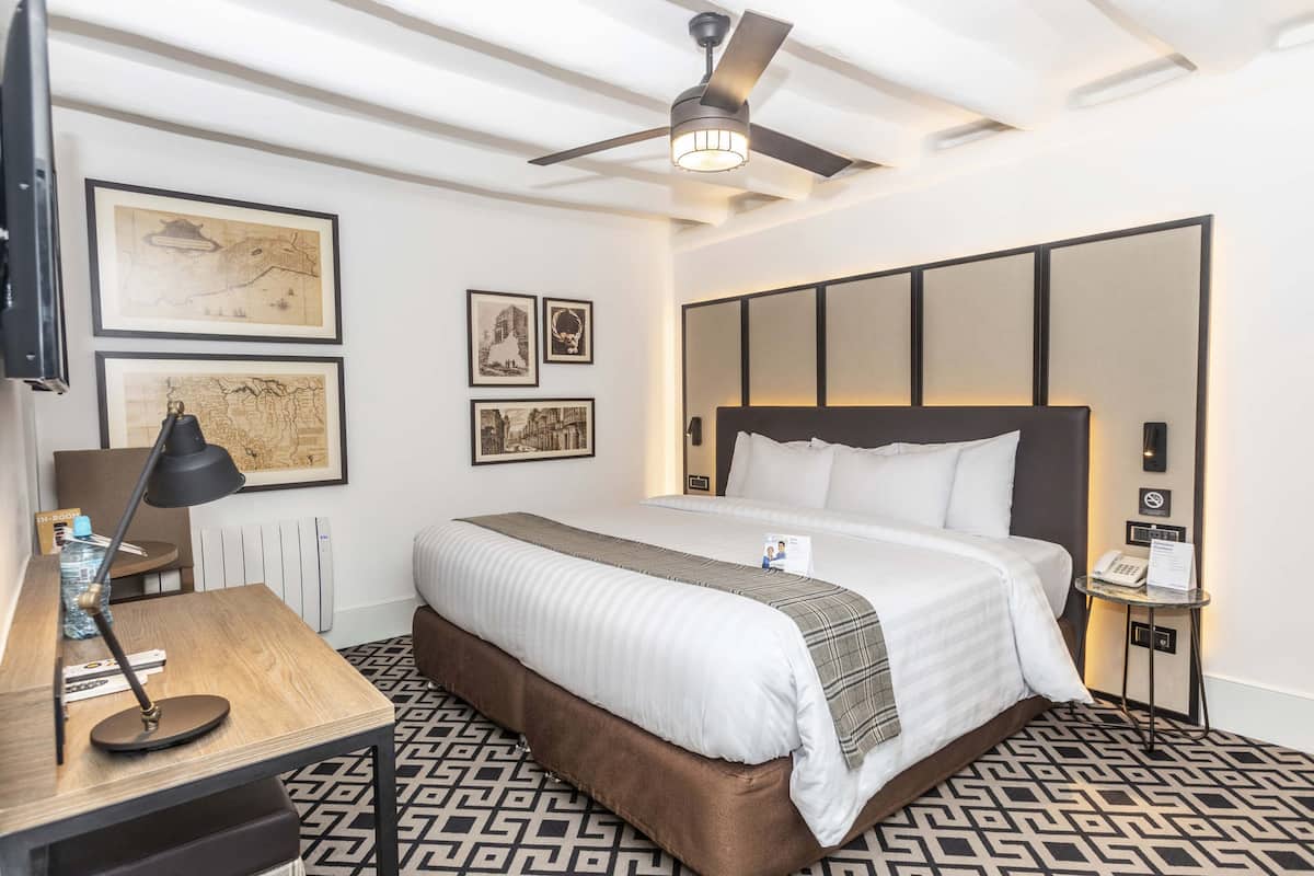 Quarto de hotel com uma cama grande, uma escrivaninha com luminária, um ventilador de teto e quadros nas paredes.