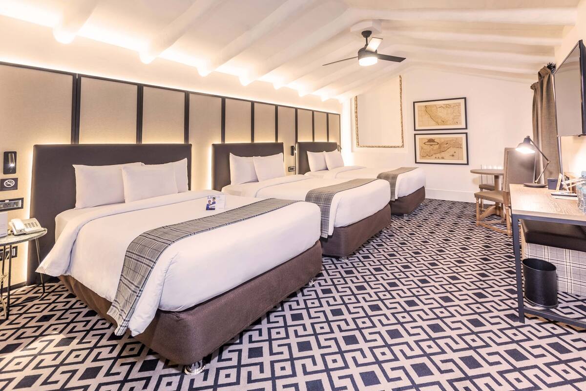 Quarto de hotel com três camas, uma escrivaninha, uma cadeira, telefone e ventoinha no teto.