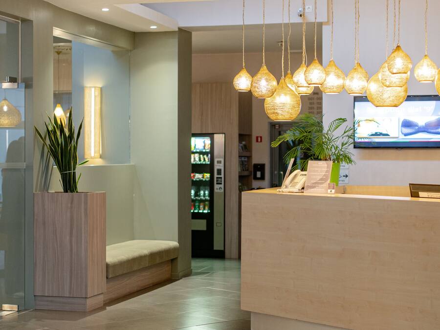 Uma área de recepção moderna com um balcão de madeira, luminárias pendentes, uma planta em vaso, um frigorífico com bebidas e uma televisão montada na parede.