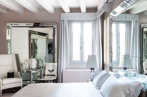 Quarto de hotel com uma cama grande, uma escrivaninha com cadeira, uma mesinha com um vaso de flores e um espelho amplo.