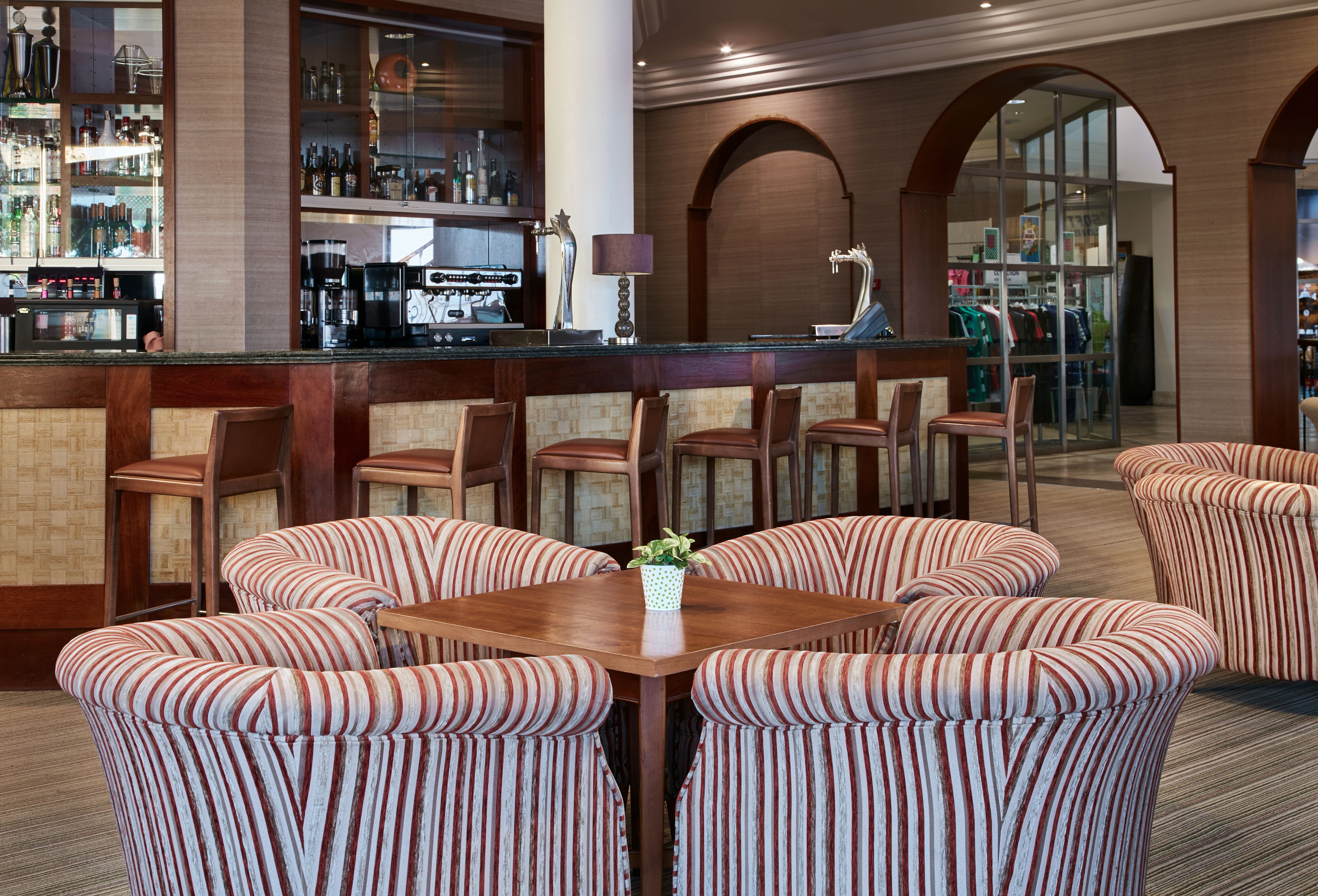 Uma área de bar com cadeiras estofadas listradas, balcão de madeira e vista para uma loja de roupas ao fundo.