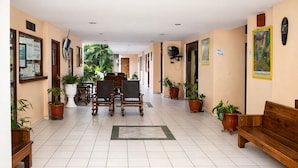 Um saguão de hotel com piso de azulejos, móveis de madeira, plantas em vasos e uma televisão presa à parede.