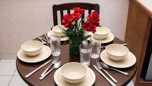 Mesa de jantar posta com pratos, tigelas e talheres brancos, acompanhados por rosas vermelhas em um vaso verde.