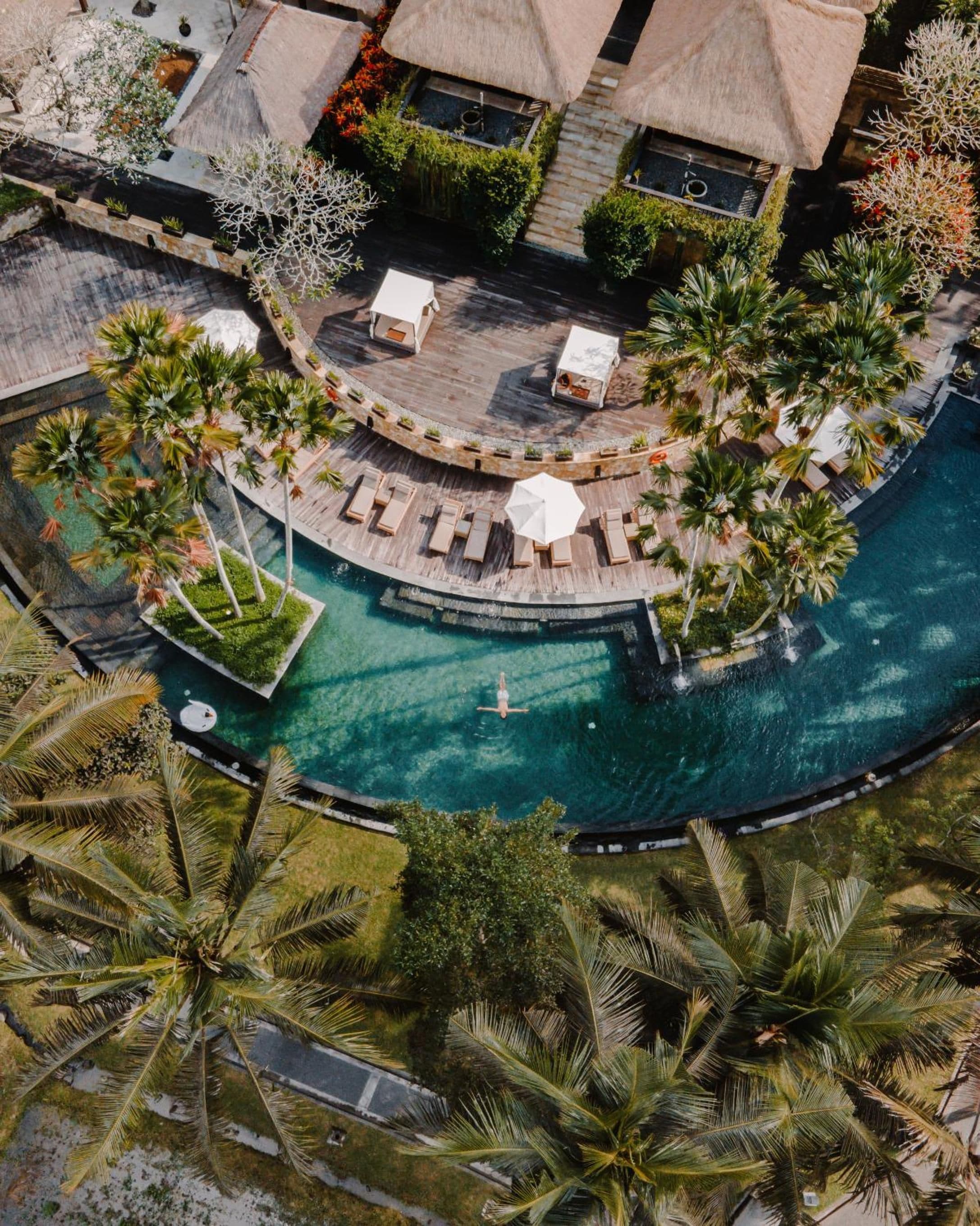 Vista aérea de um resort com piscina circular, áreas de descanso e palmeiras.
