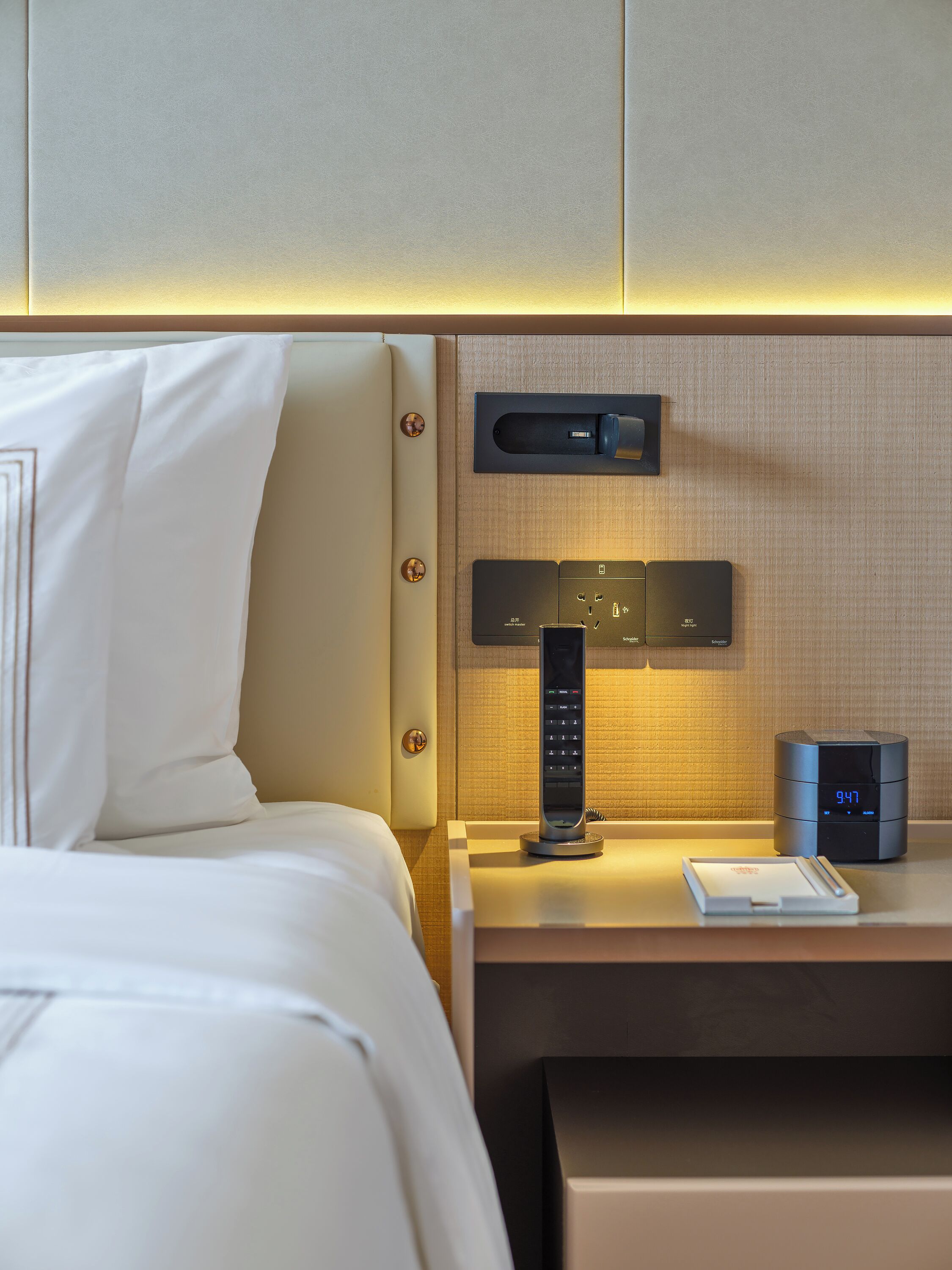 Quarto de hotel com uma cama bem arrumada, uma mesa de cabeceira com relógio digital e um painel de controle fixado na parede.