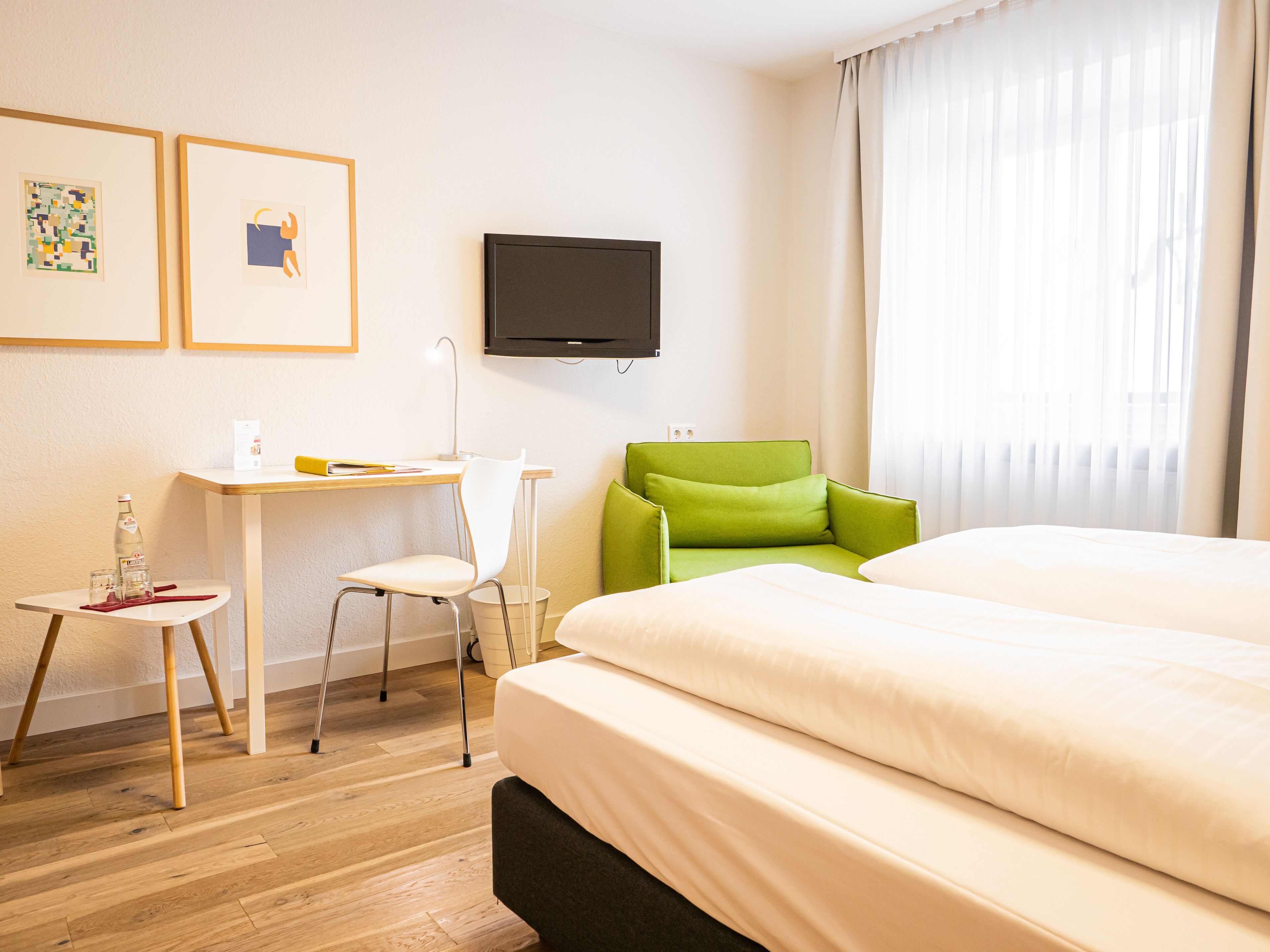 Quarto de hotel com cama, escrivaninha, cadeira, televisão e obras de arte na parede.