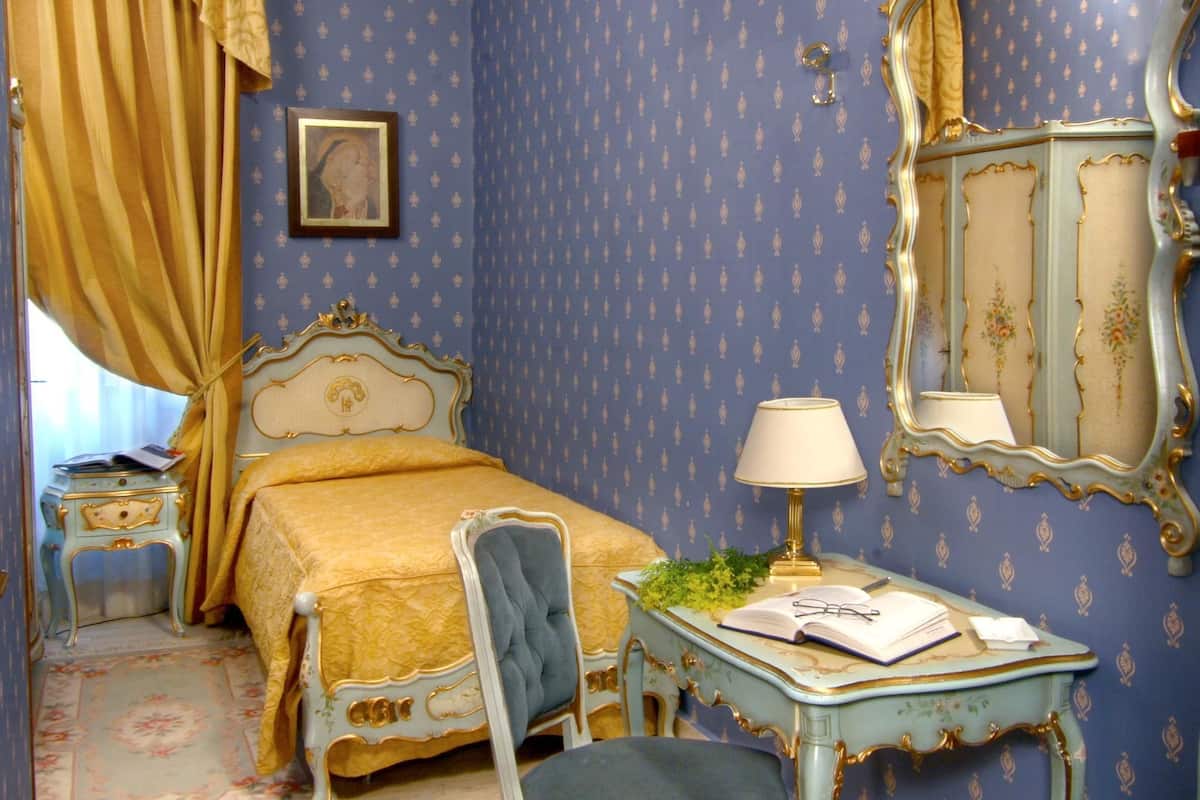 Quarto com cama, escrivaninha e cadeira. Há um espelho na parede e um lustre sobre a cama.
