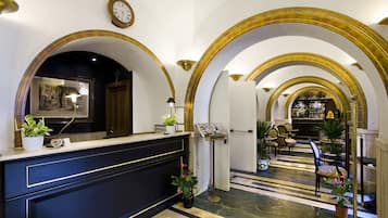 Um saguão de hotel com balcão de recepção, obras de arte emolduradas, plantas em vasos e um bar ao fundo.