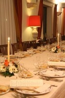 Uma longa mesa de jantar posta para uma refeição formal, com velas, arranjos florais e um menu.