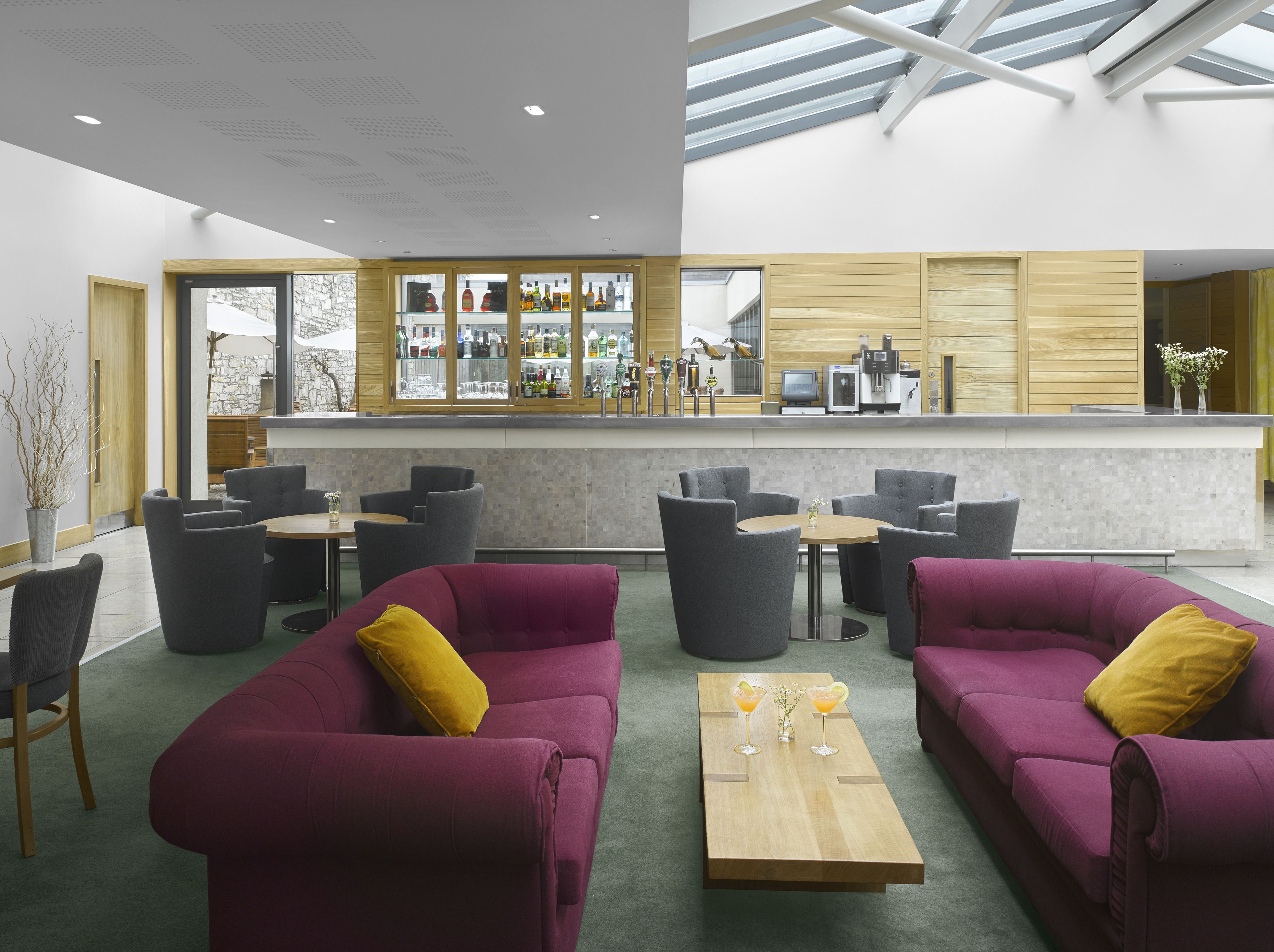 Um lobby moderno com sofás roxos, uma mesa de centro de madeira e uma área de bar com garrafas e uma televisão.