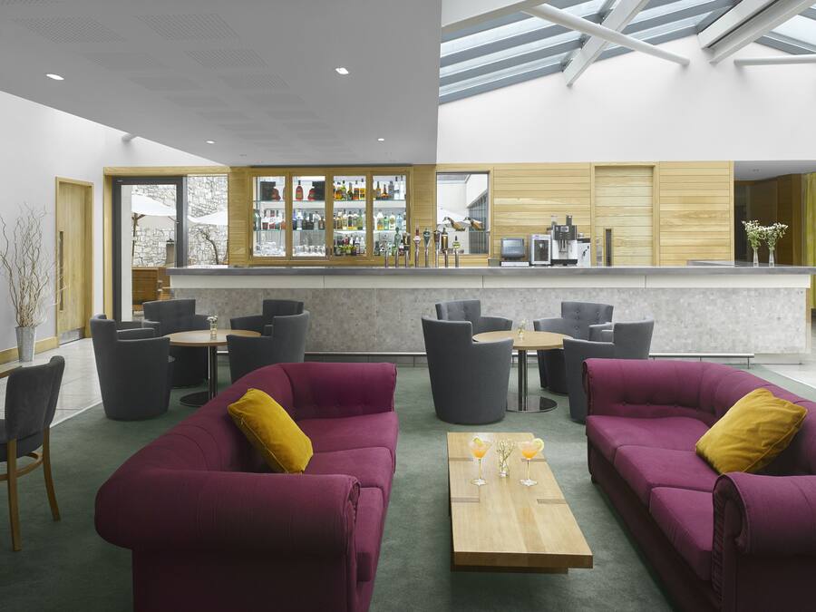Um lobby moderno com sofás roxos, uma mesa de centro de madeira e uma área de bar com garrafas e uma televisão.