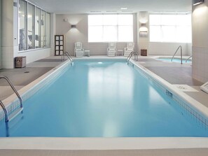 Uma grande piscina interna com água azul cristalina, rodeada por cadeiras brancas, e uma área separada com hidromassagem.