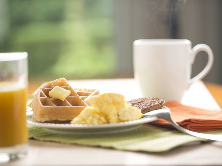 Uma mesa de café da manhã com waffles, ovos mexidos, torradas, um copo de suco de laranja e uma caneca branca, tudo sobre um jogo americano verde.