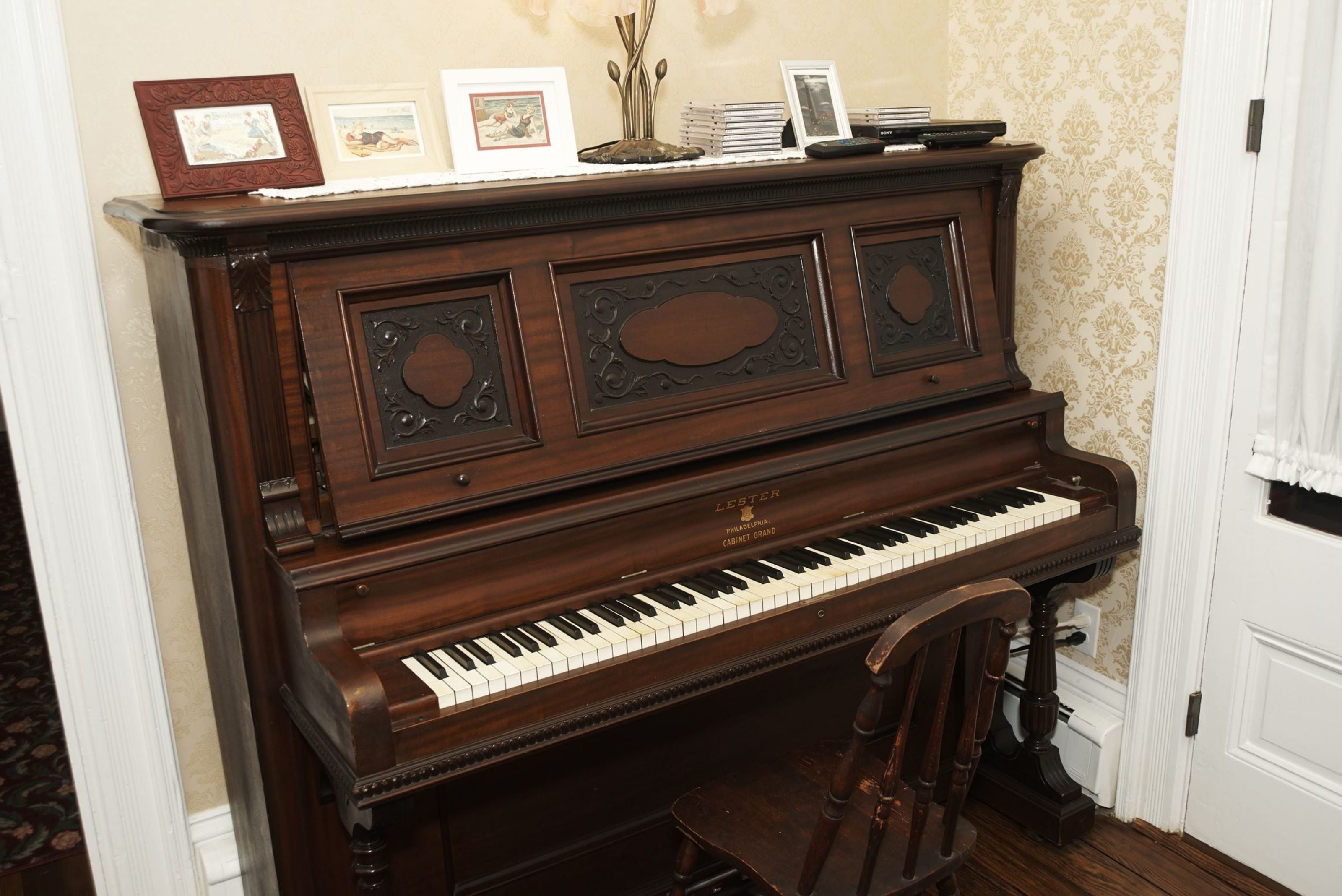 Um piano de cauda com acabamento em madeira e painéis decorativos, colocado numa sala com papel de parede estampado e uma porta branca.