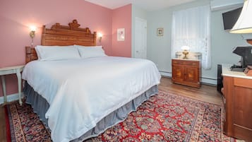 Quarto com uma cama grande, cabeceira de madeira, mesa de cabeceira, escrivaninha, cadeira, luminária de mesa, televisão e tapete estampado.