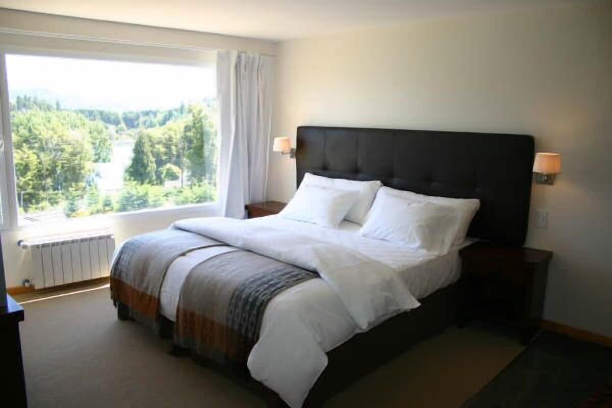 Quarto com uma cama grande, duas luminárias de cabeceira e vista para uma floresta pela janela.