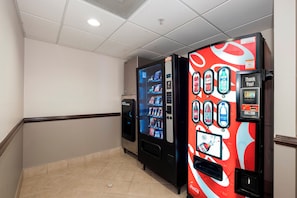 Corredor com máquinas de venda automática, incluindo uma da marca Coca-Cola, e um bebedouro.