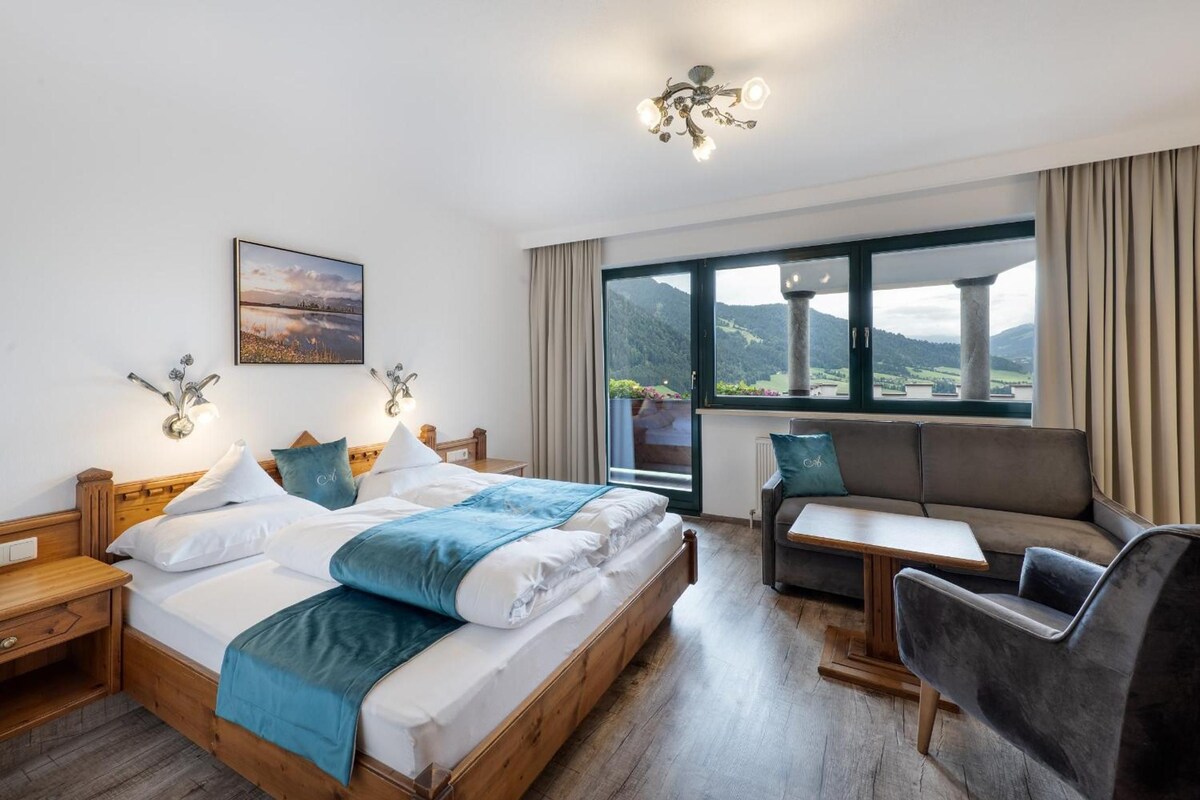 Quarto de hotel com cama, sofá, mesinha e grande janela com vista para montanhas.