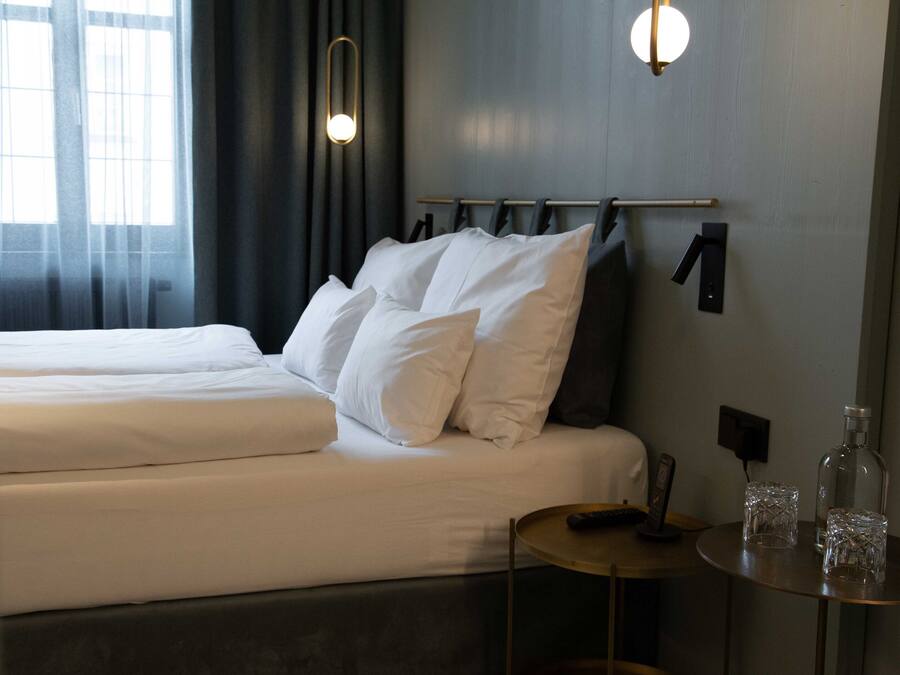 Quarto de hotel com duas camas, uma mesinha de cabeceira com um controle remoto e uma pequena mesa com dois copos.