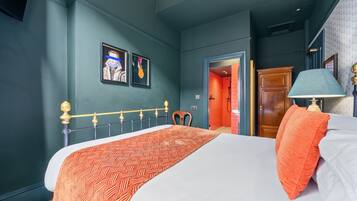 Uma cama bem arrumada com um cobre-leito laranja estampado, uma cabeceira de madeira e um único travesseiro laranja. Há duas quadros na parede acima da cama.