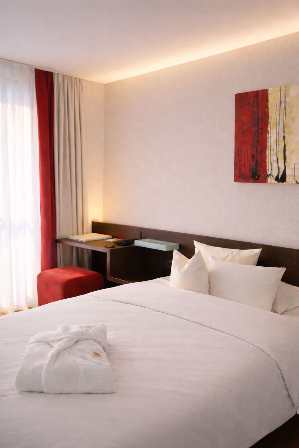 Quarto de hotel com uma cama grande, uma escrivaninha, um banquinho vermelho e um quadro na parede.