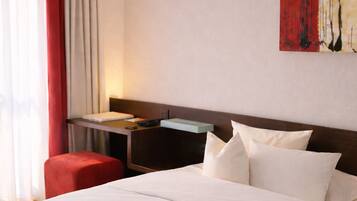 Quarto de hotel com uma cama grande, uma escrivaninha, um banquinho vermelho e um quadro na parede.