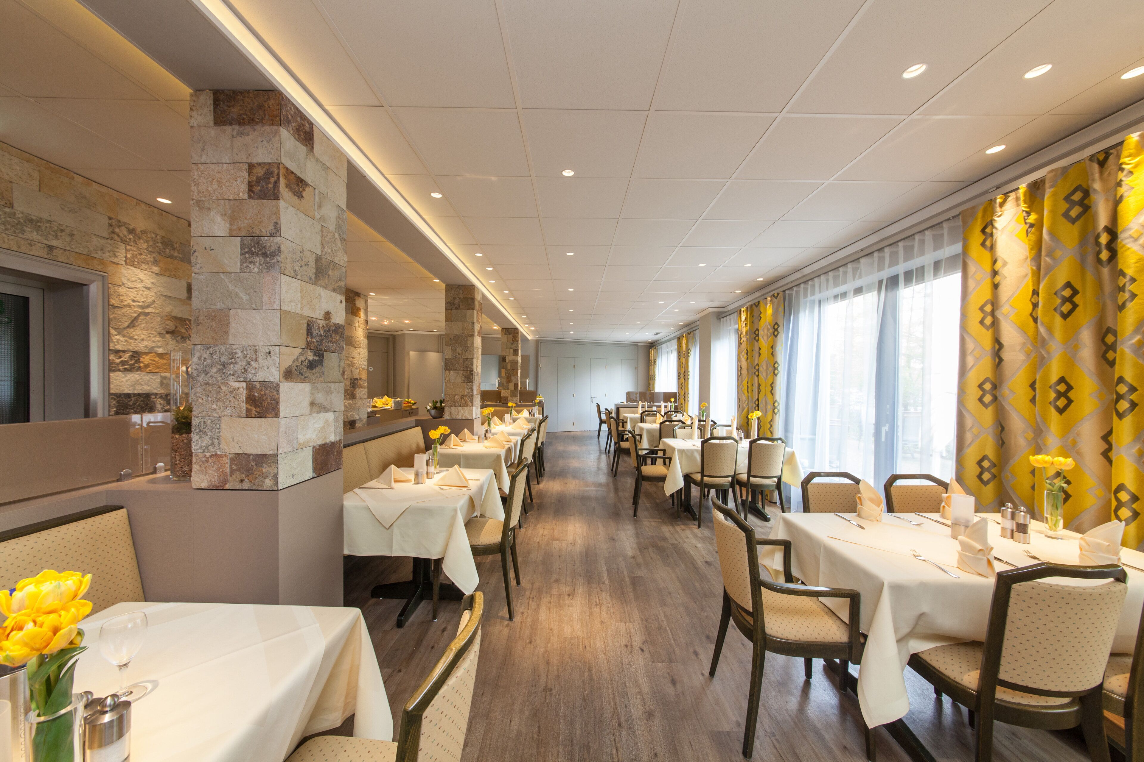 Um restaurante com várias mesas, pilares de pedra e cortinas estampadas em amarelo.