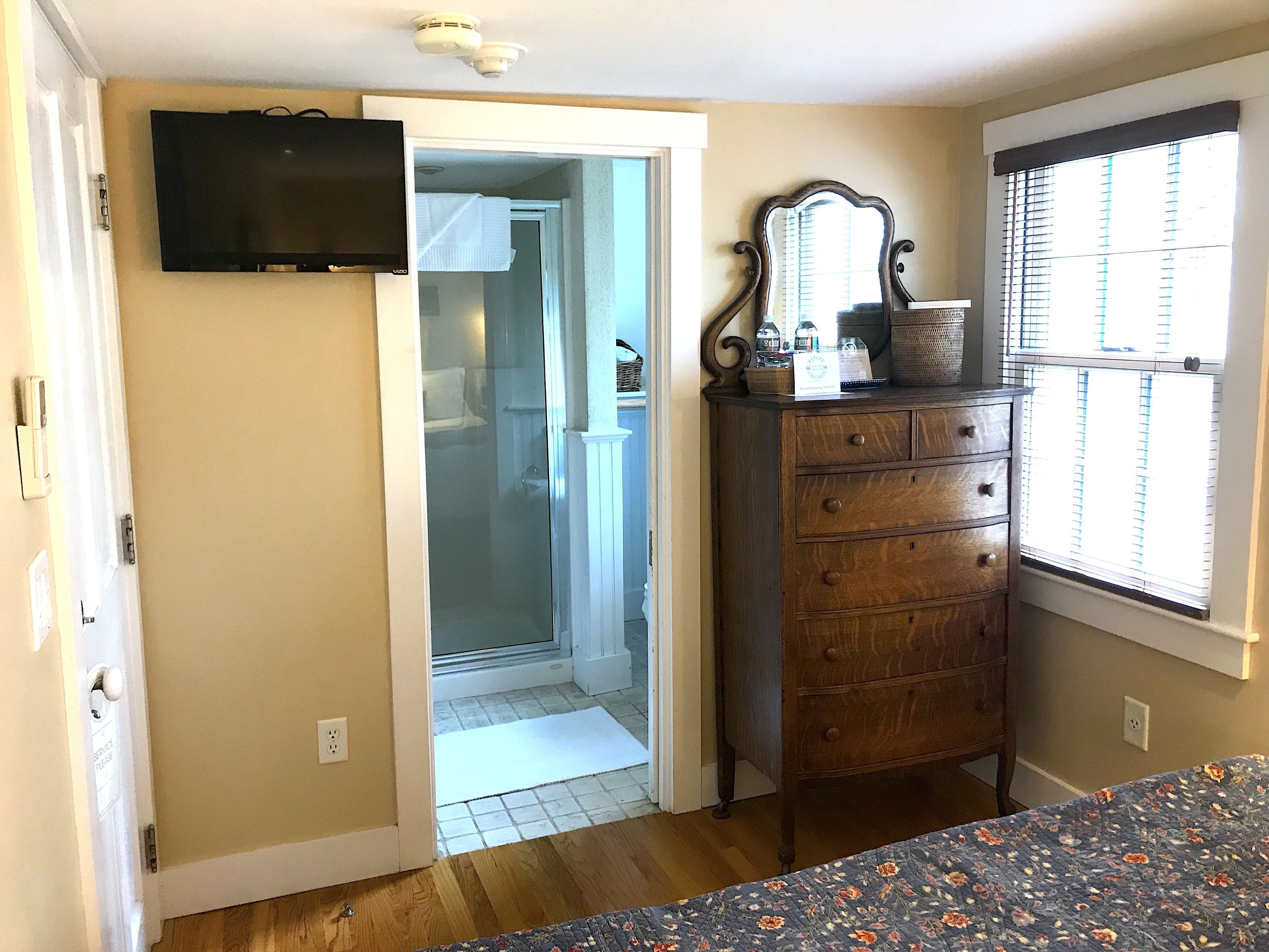 Quarto com cômoda de madeira, televisão fixada na parede, janela com persianas e porta de acesso a um banheiro.