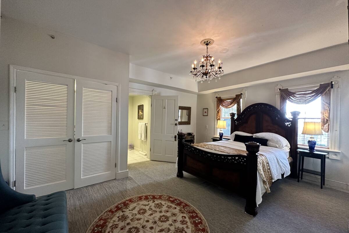 Um quarto com uma cama grande, um candelabro, uma janela com cortinas e uma porta com venezianas.