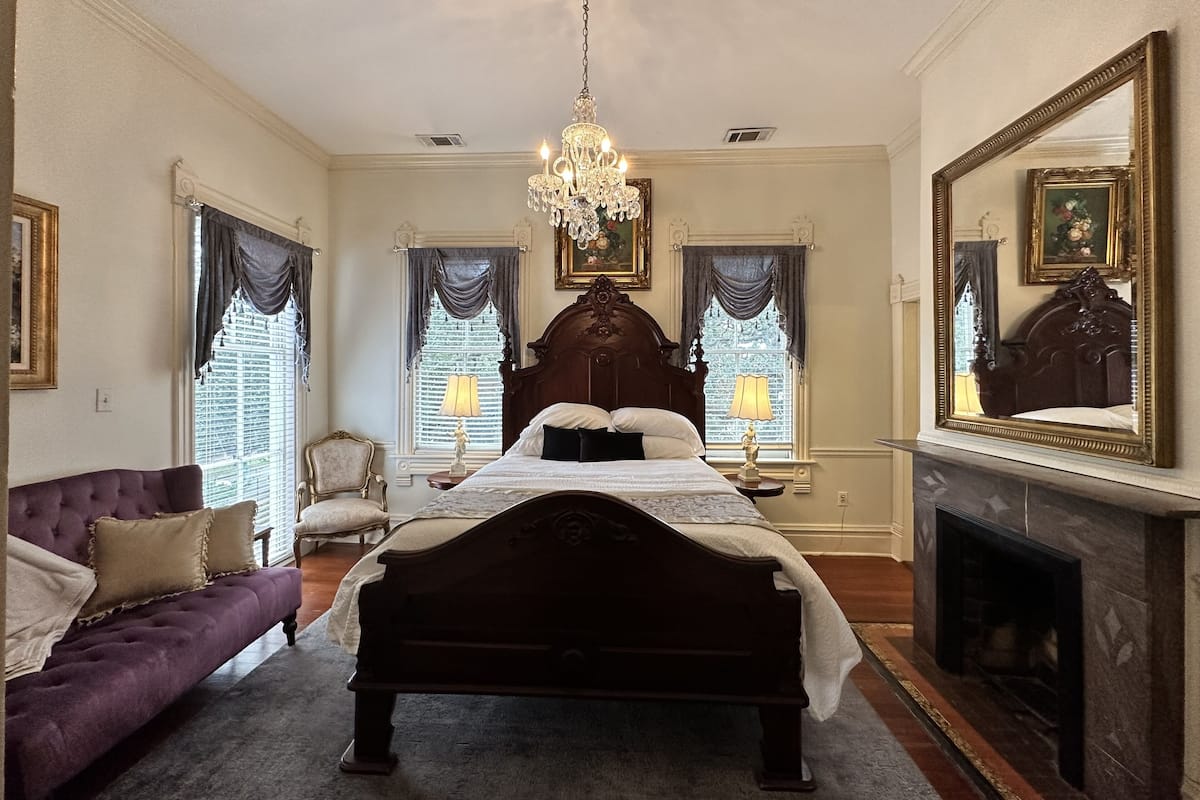 Quarto com cama grande, lustre, lareira e sofá roxo.