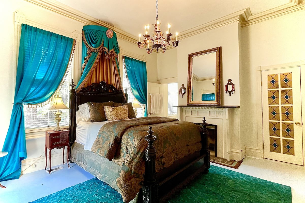 Quarto com cama de dossel, lustre, lareira e um espelho grande.