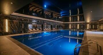 Uma grande piscina interior com paredes e teto de mármore, rodeada por espreguiçadeiras e uma área de lazer junto à piscina.