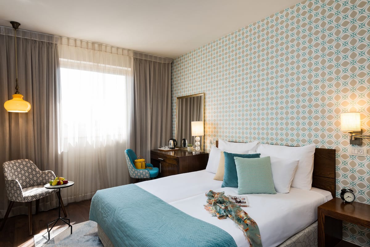 Quarto de hotel com cama, escrivaninha, cadeira, abajur e relógio.