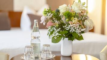 Quarto de hotel com cama, mesa de cabeceira com garrafa e copos, e um vaso com flores.