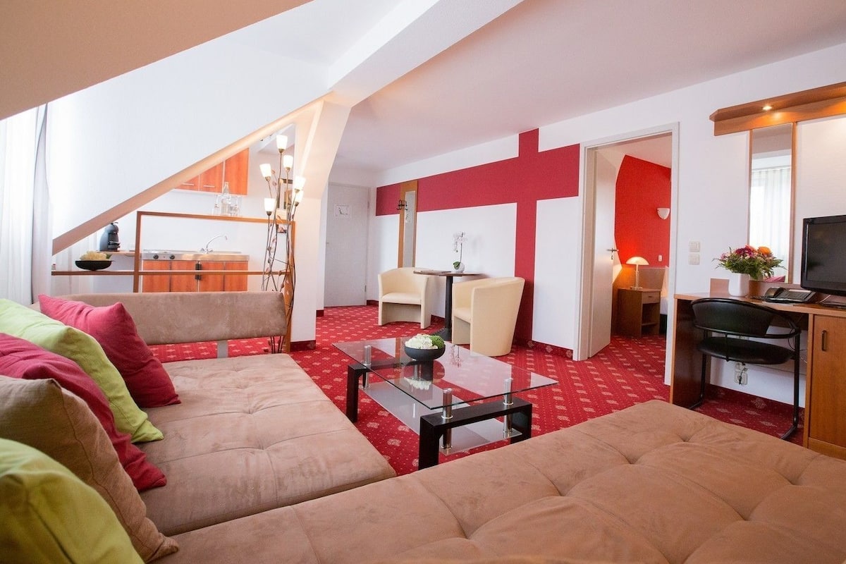 Quarto de hotel moderno com teto estilo loft, área de estar aconchegante com sofá e banquinho, mesa de jantar com cadeiras, pequena cozinha e televisão.