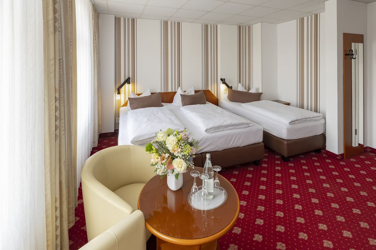 Quarto de hotel com duas camas, uma mesa de madeira com um vaso de flores e chão coberto por um tapete vermelho.