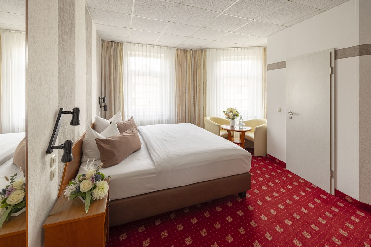 Quarto de hotel com uma cama grande, uma escrivaninha e uma cadeira. Há um tapete vermelho e uma janela com cortinas.