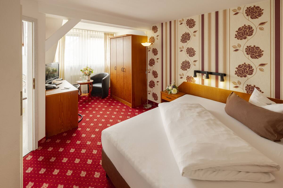 Quarto de hotel com cama, escrivaninha, cadeira, guarda-roupa e janela com cortinas.