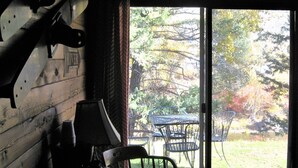 Interior de cabana com parede de madeira, mesa, cadeiras e porta de vidro deslizante que dá para área externa com cadeiras e mesa.