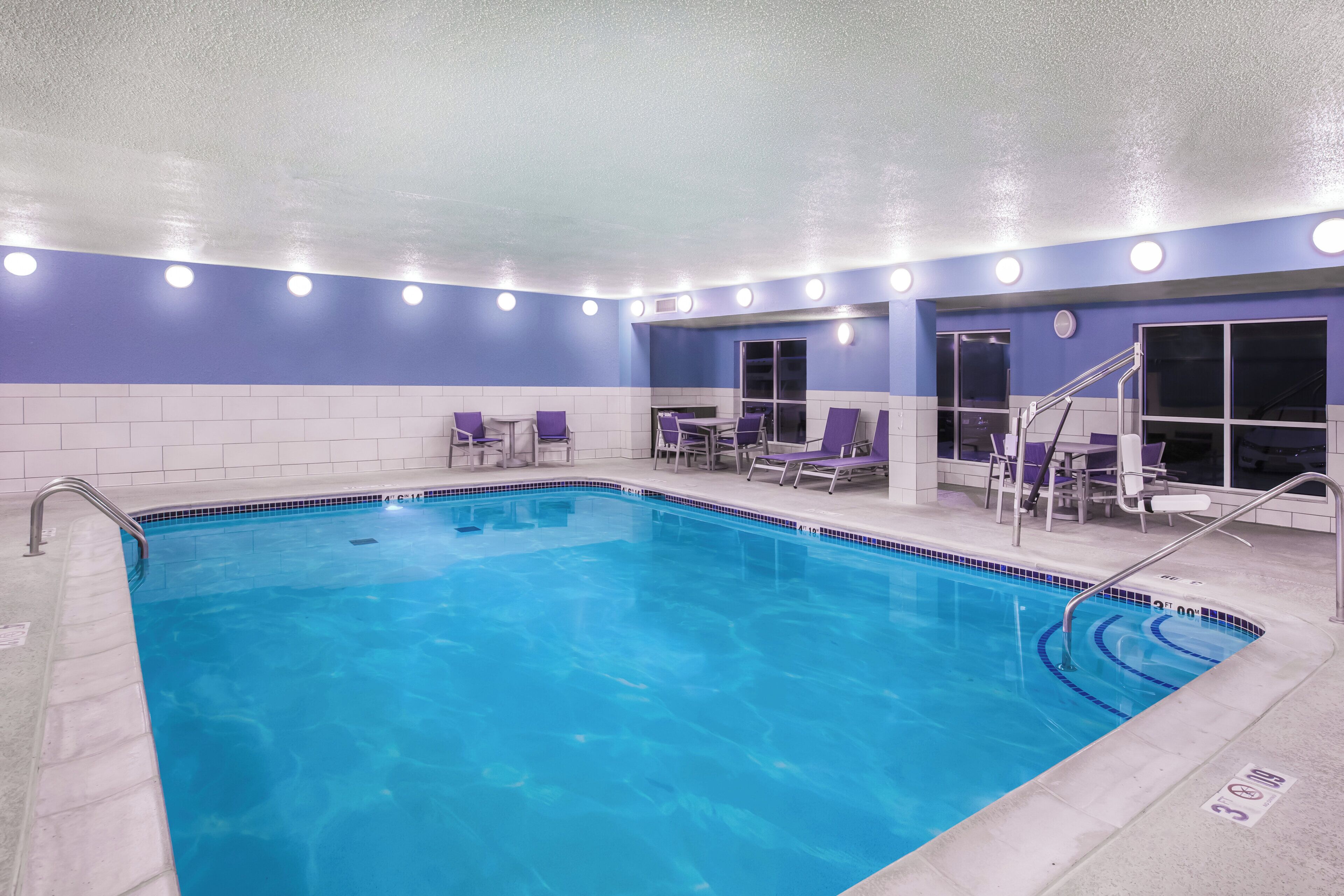 Piscina interna com água azul cristalina, rodeada por cadeiras de descanso roxas e um revestimento de azulejos.