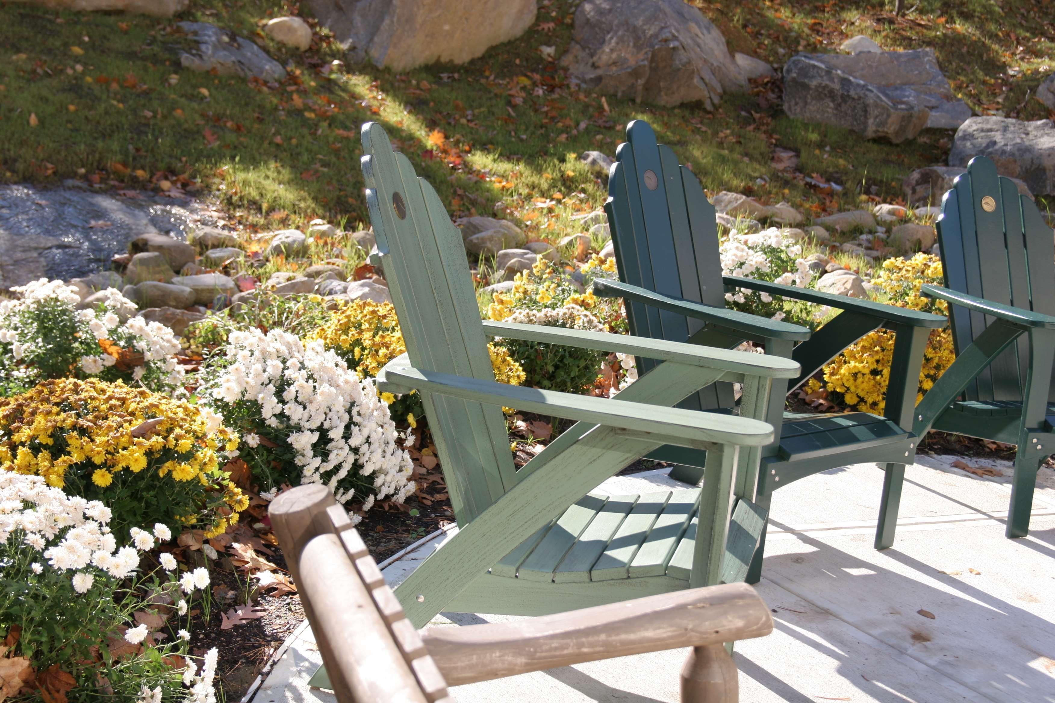 Um deck de madeira com cadeiras Adirondack e um banco, cercado por um jardim com diversas flores.