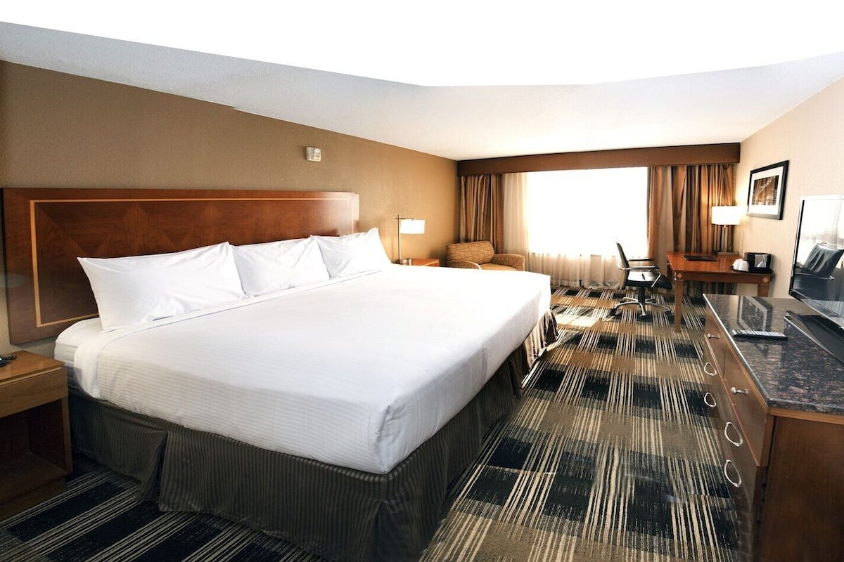 Quarto de hotel com uma cama grande, uma escrivaninha com televisão de tela plana, uma cadeira e uma luminária de mesa.
