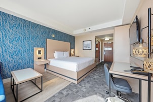 Quarto de hotel com cama, mesas de cabeceira, escrivaninha, cadeira e televisão.