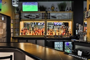 Uma área de bar com prateleiras cheias de garrafas de bebidas, uma televisão exibindo um evento esportivo e um balcão com um caixa registradora.