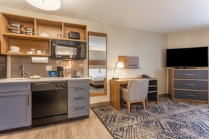 Quarto de hotel compacto com cozinha compacta, uma cama, uma escrivaninha com cadeira, televisão e pequeno armário.