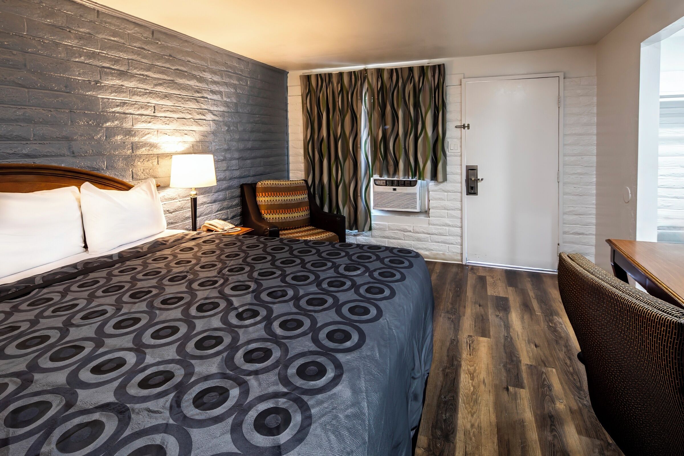 Quarto de hotel com uma cama grande, mesa de cabeceira, cadeira, ar condicionado fixado na parede e uma porta com cortinas.