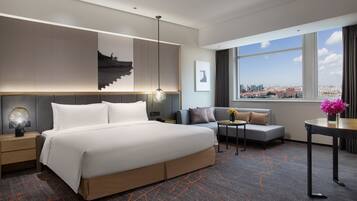 Quarto de hotel moderno com uma cama grande, um sofá, uma pequena mesa com flores e vista para a cidade através de grandes janelas.